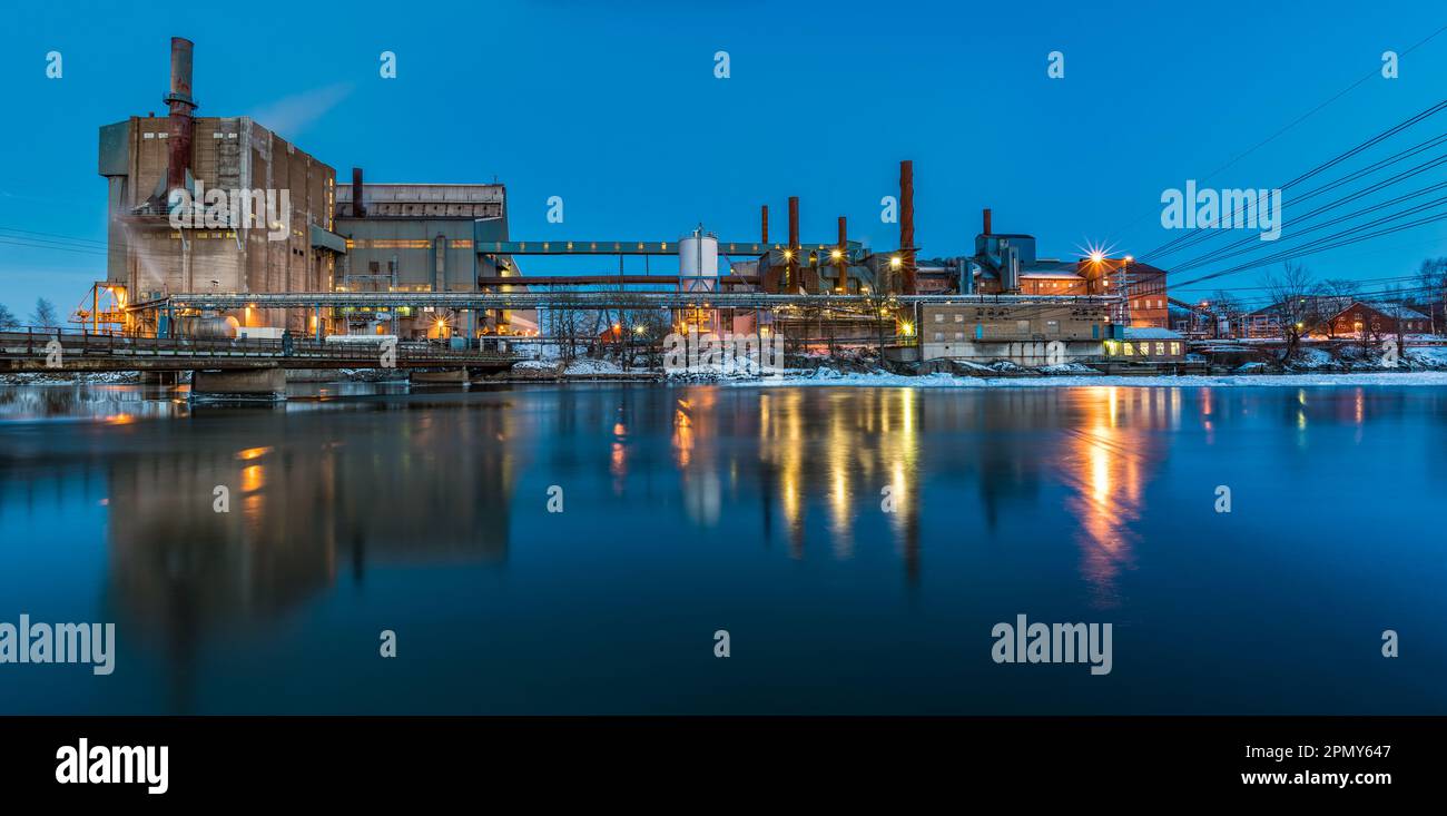 At dusk, the illuminated factory casts a reflection onto the river ...