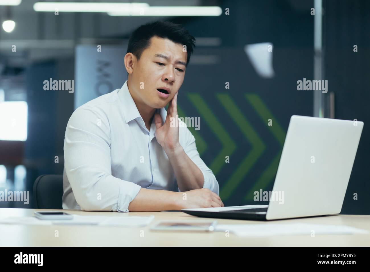 Toothache at work. A young Asian man is sitting at the desk in the ...