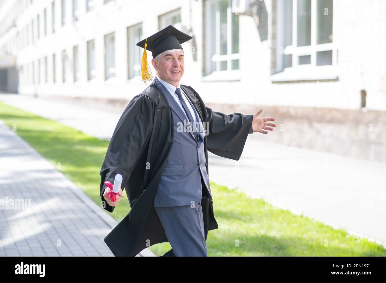 An old man in a graduation gown walks outdoors and holds a diploma ...