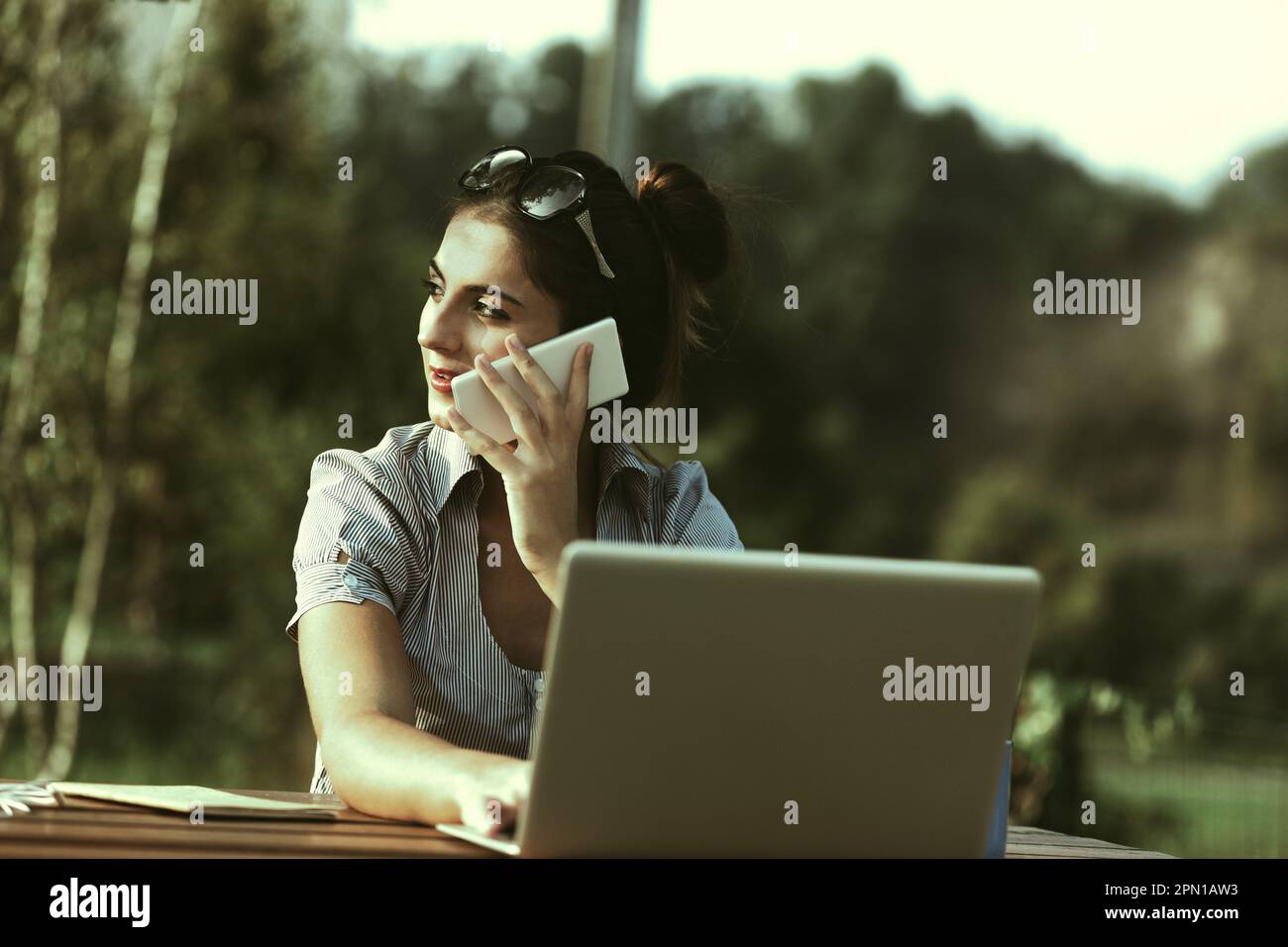 A woman facing left talks on her smartphone while using a laptop ...