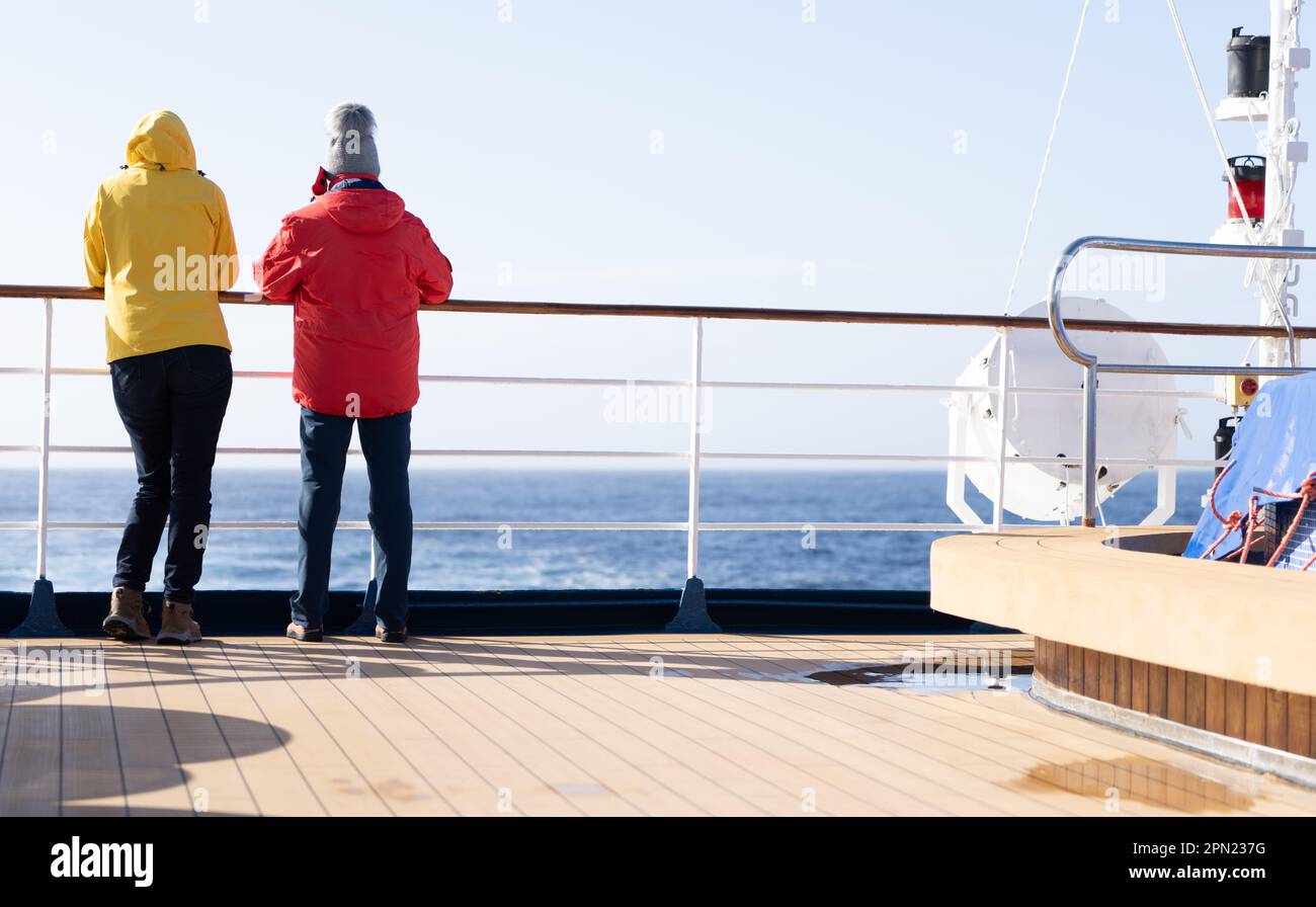2 cruise passengers stands at the railing at the stern of a cruise ship ...