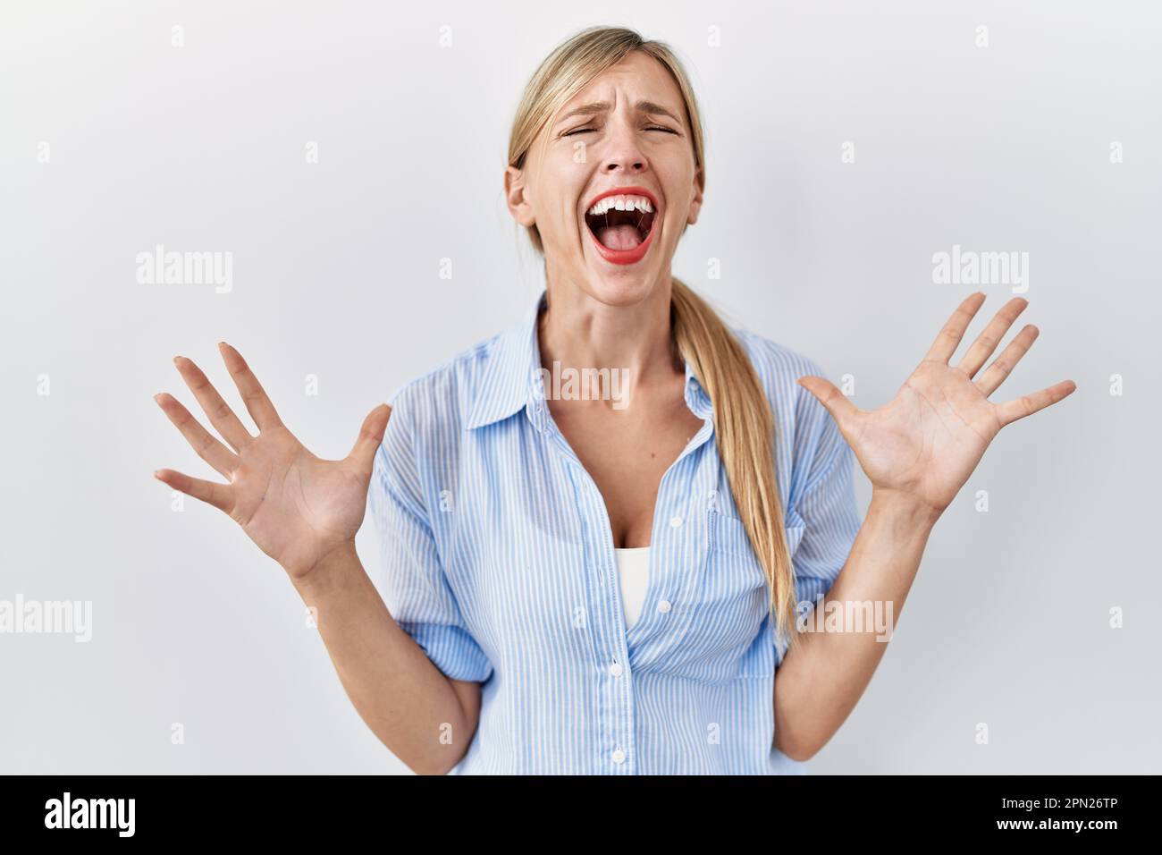 Beautiful blonde woman standing over white background crazy and mad ...