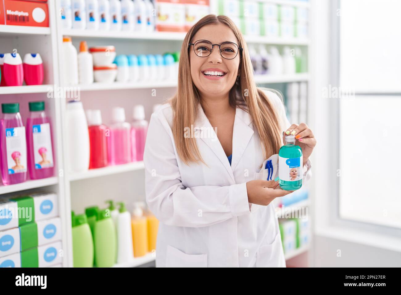 Young blonde woman working at pharmacy drugstore holding syrup smiling ...
