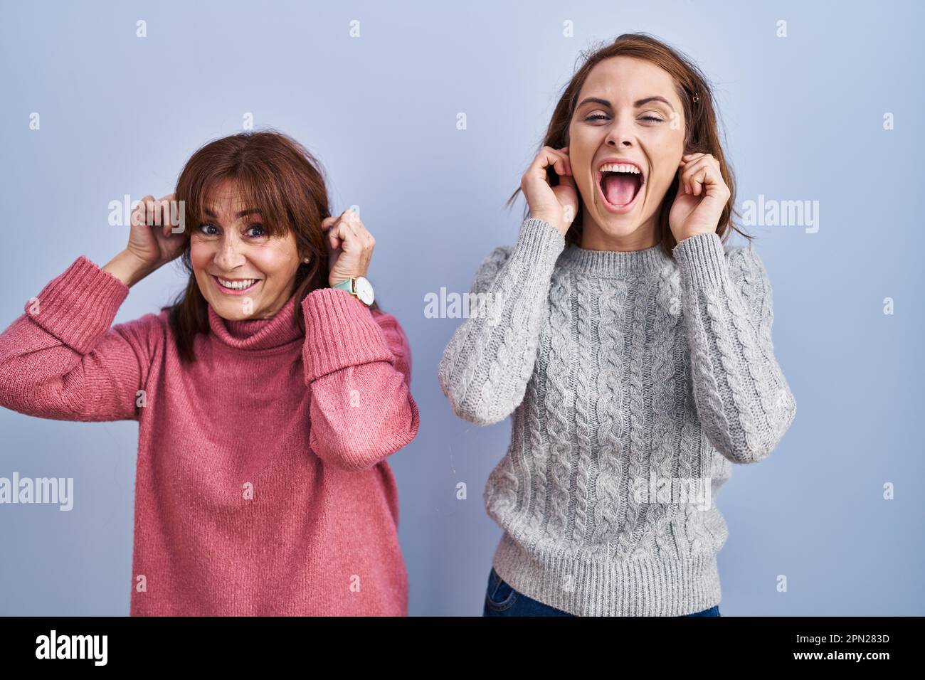 Mother and daughter standing over blue background smiling pulling ears ...