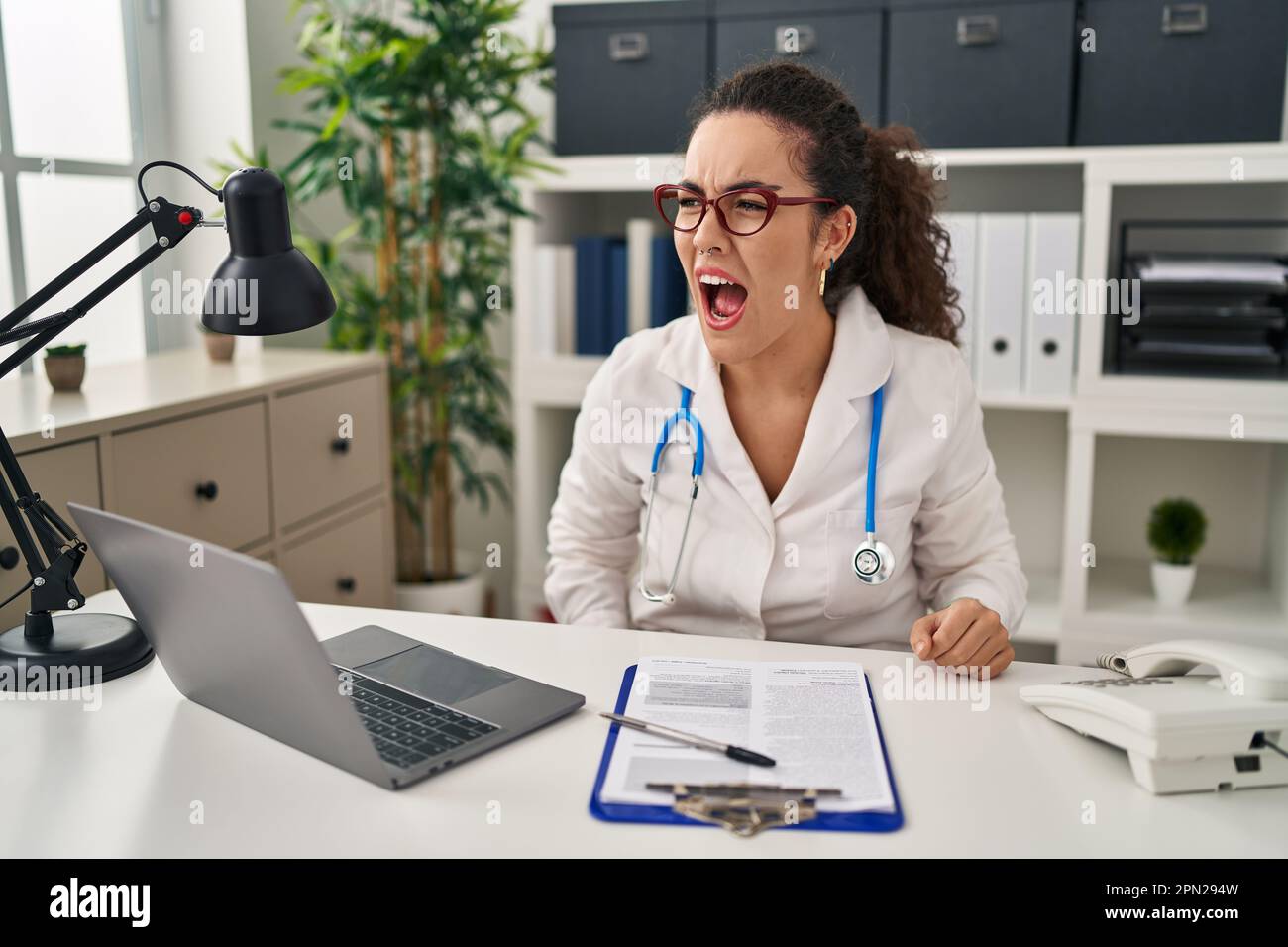 Young hispanic woman wearing doctor uniform and stethoscope angry and ...