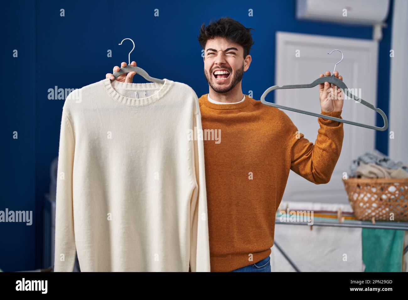 Hispanic man with beard holding sweater on hanger at laundry room angry ...