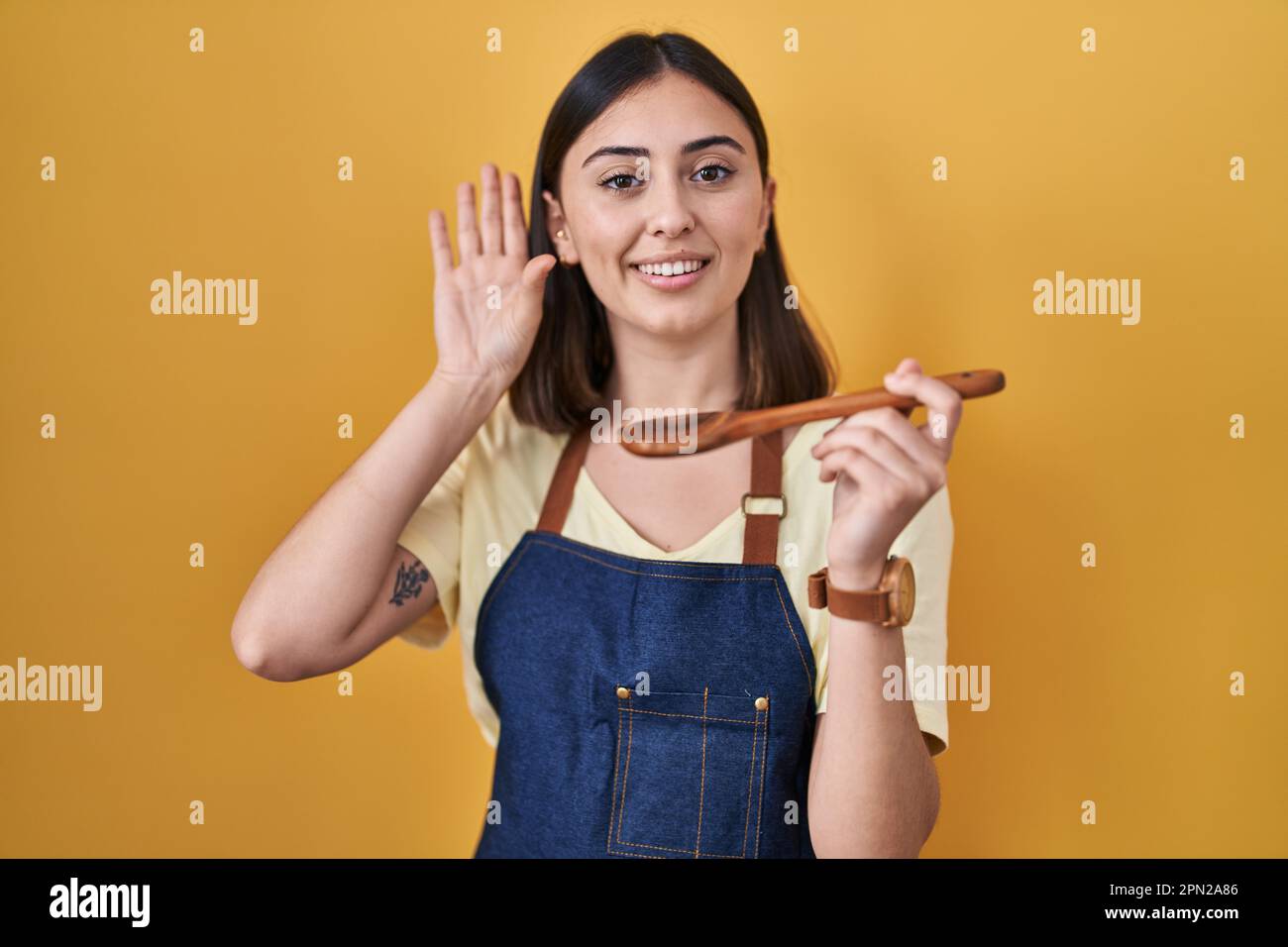Hispanic girl eating healthy wooden spoon waiving saying hello happy ...