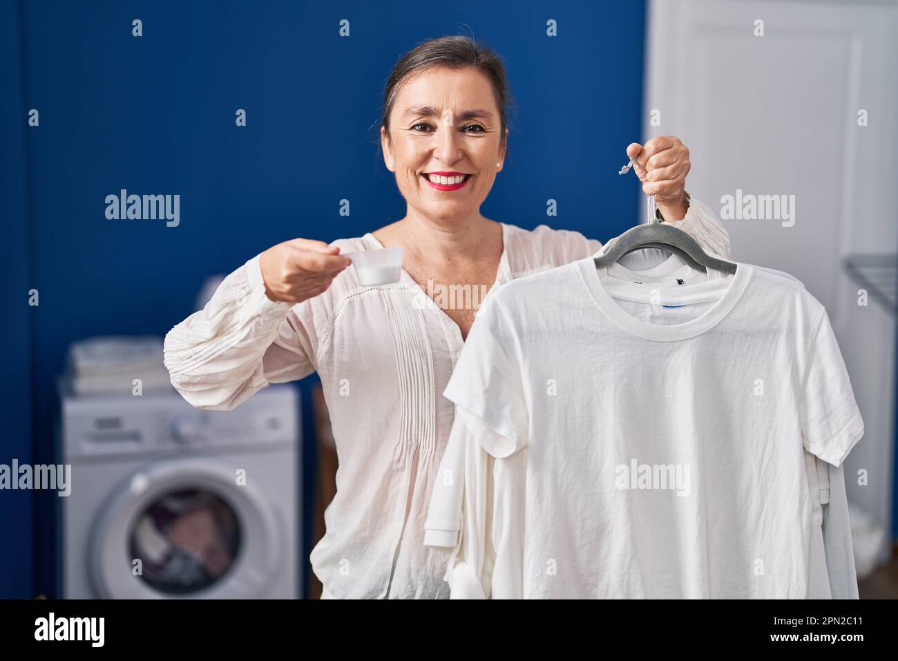 Middle age hispanic woman holding shirt on hanger and detergent powder ...