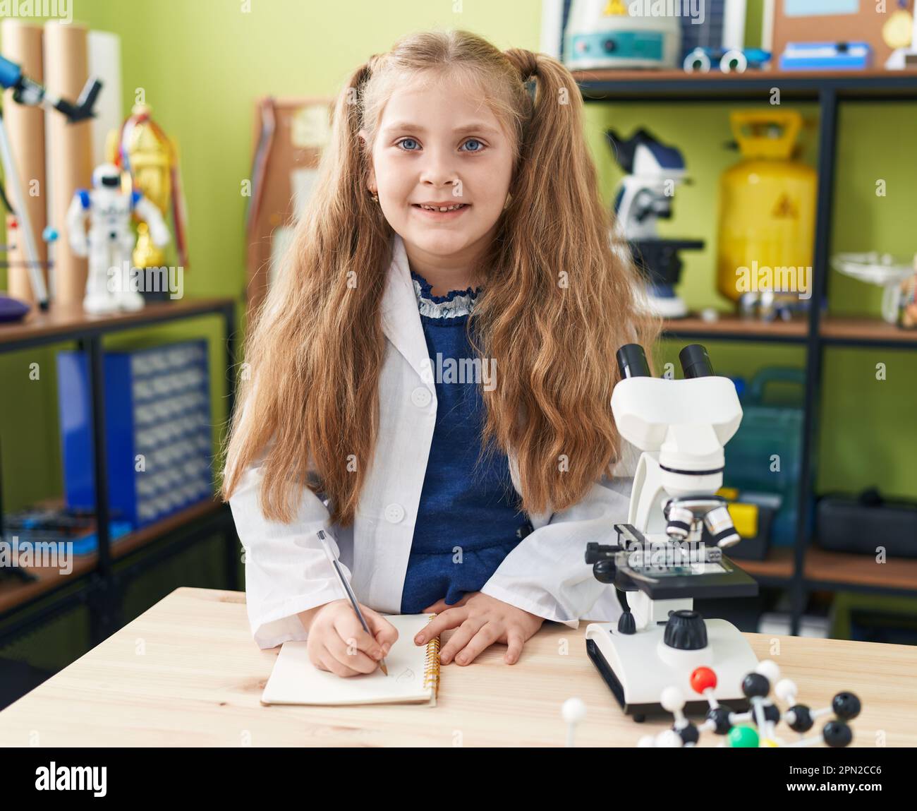 Adorable blonde girl student writing notes at laboratory classroom ...