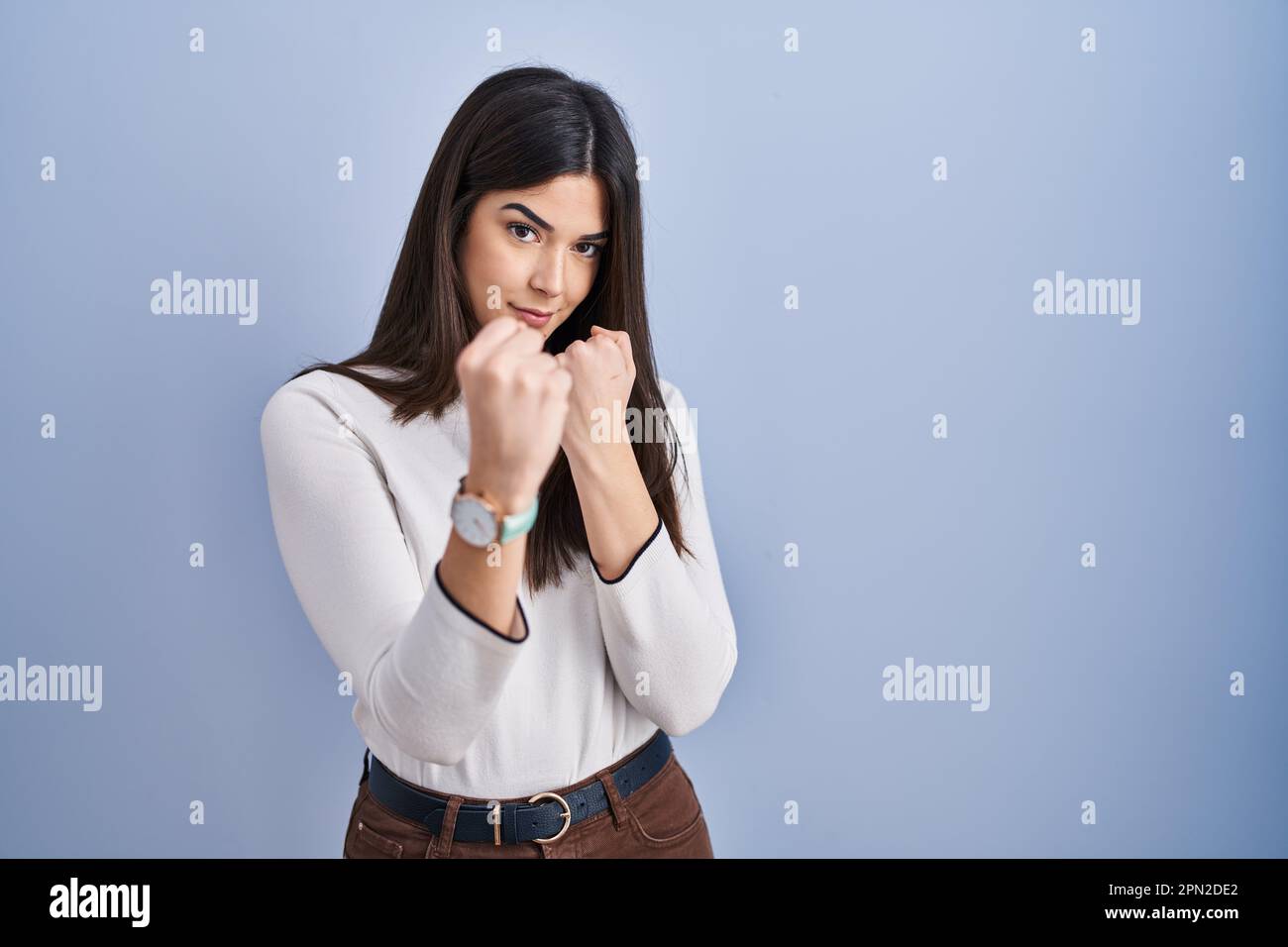 Young brunette woman standing over blue background ready to fight with ...