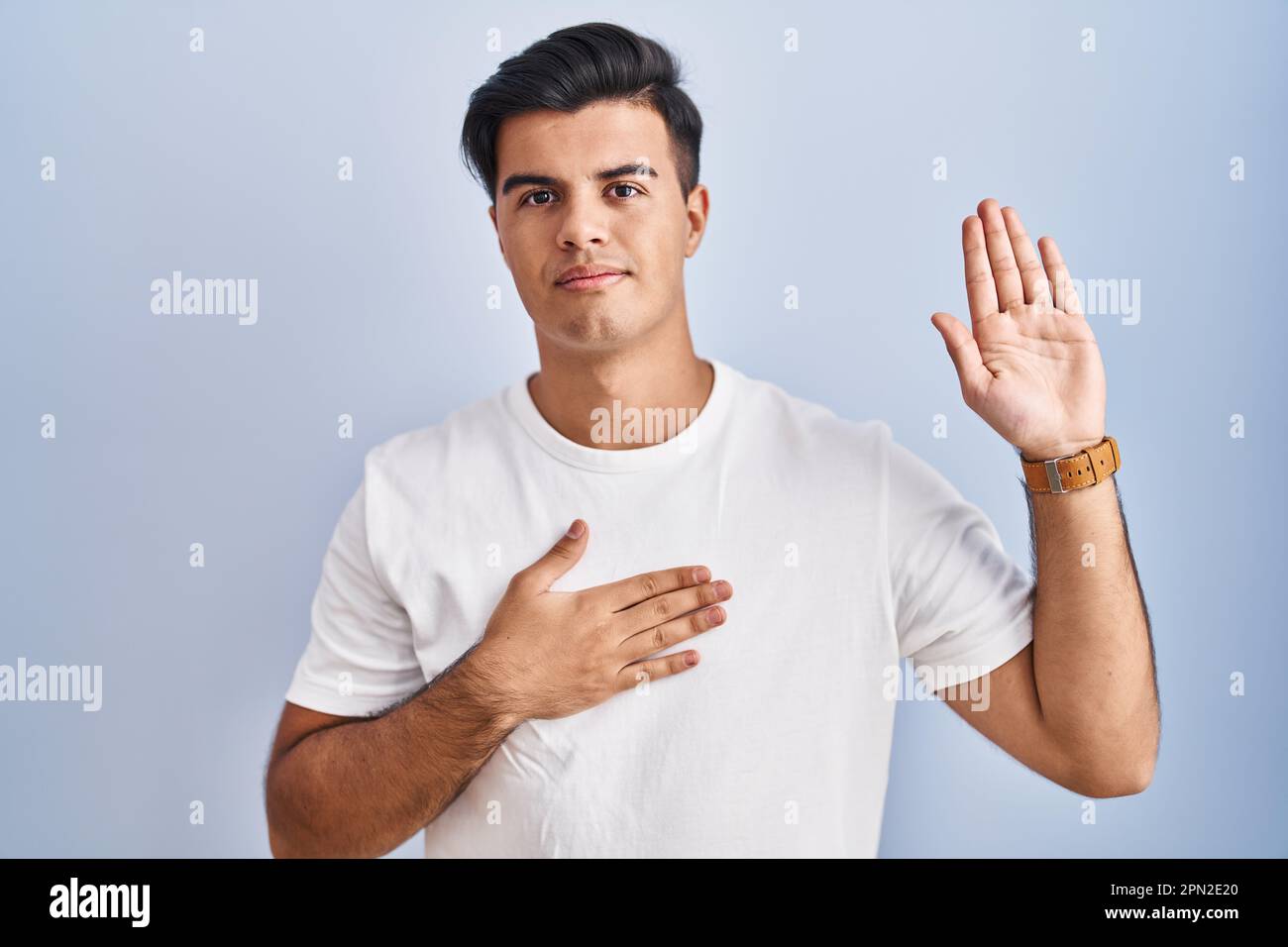 Hispanic man standing over blue background swearing with hand on chest ...