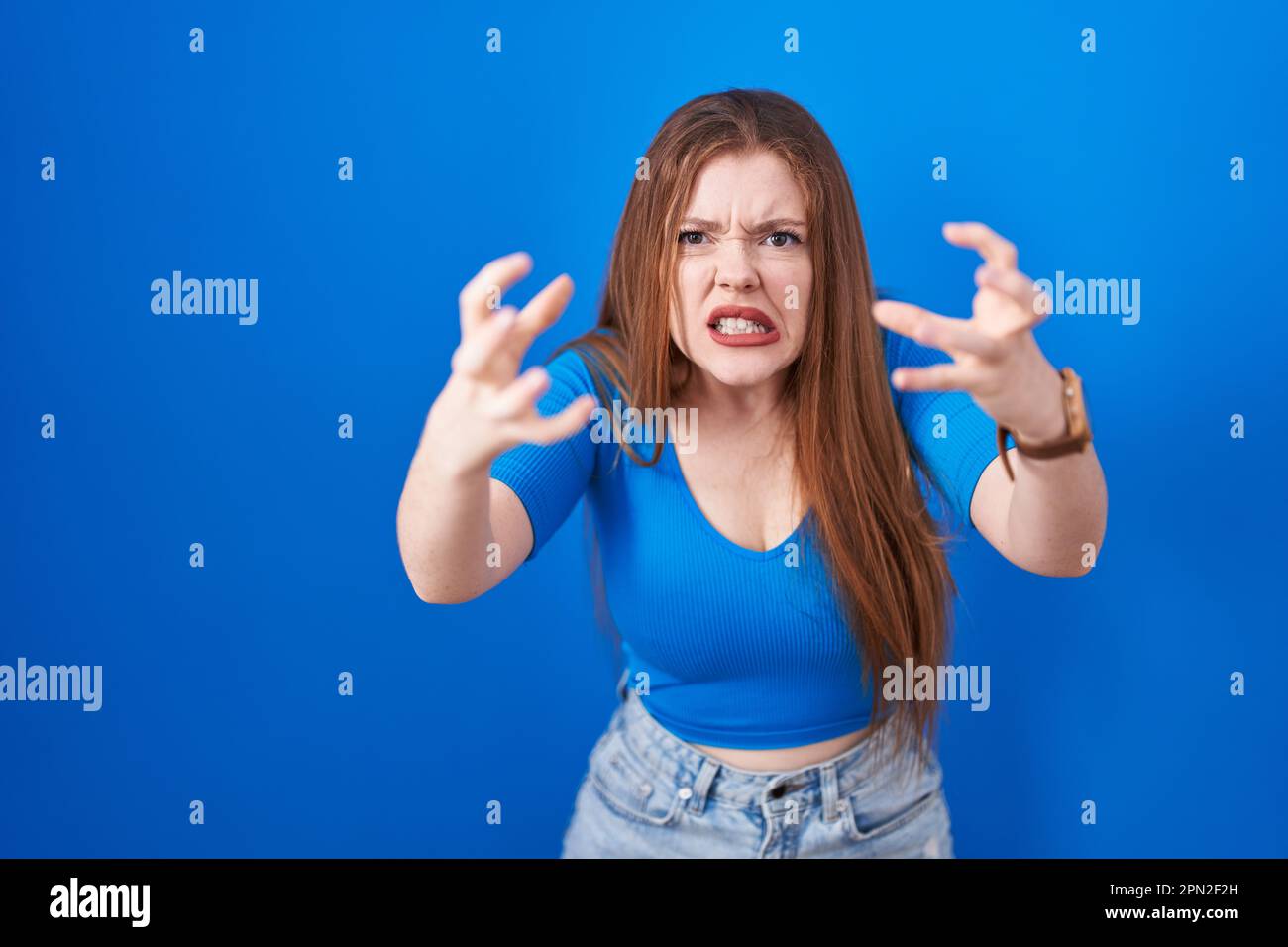 Redhead woman standing over blue background shouting frustrated with ...