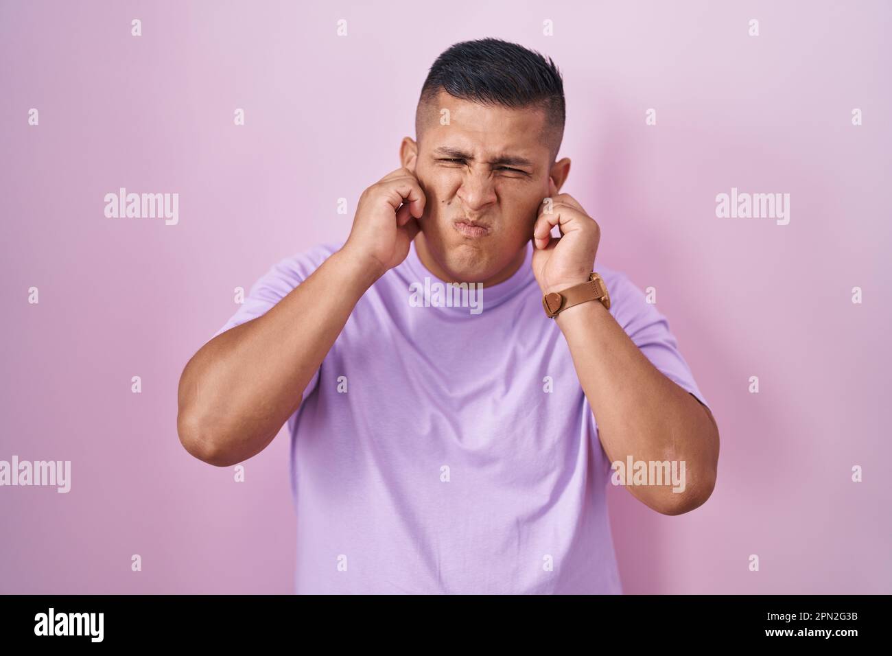 Young hispanic man standing over pink background covering ears with ...