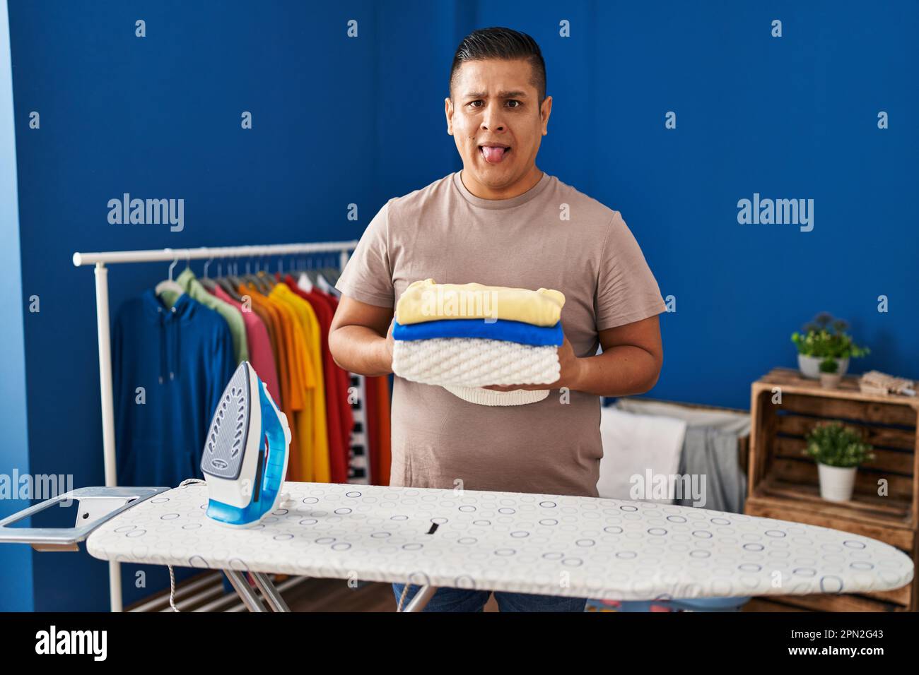 Hispanic young man holding folded laundry after ironing sticking tongue ...