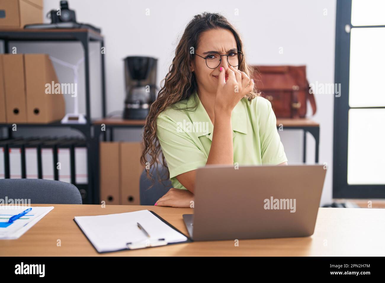 Young hispanic woman working at the office wearing glasses smelling ...