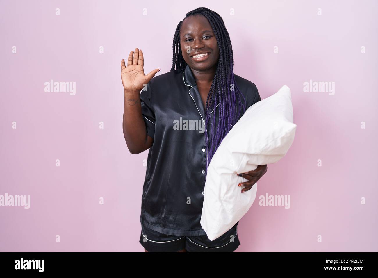 Young african woman wearing pijama hugging pillow waiving saying hello ...