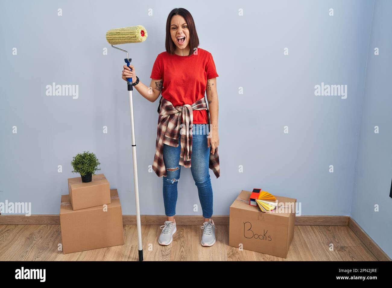 Young hispanic woman painting home walls with paint roller angry and ...