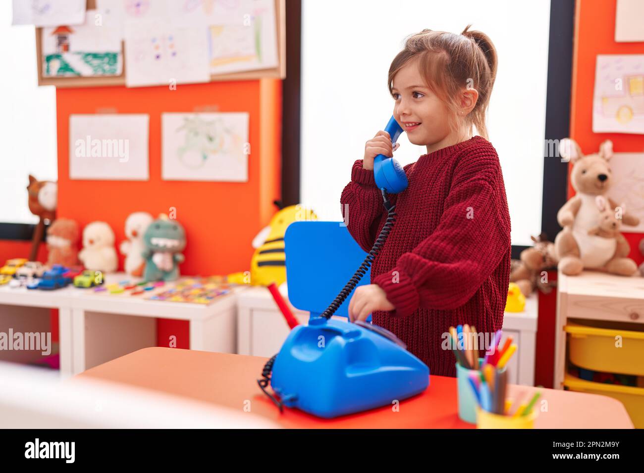 Adorable hispanic girl playing telephone toy standing at kindergarten ...
