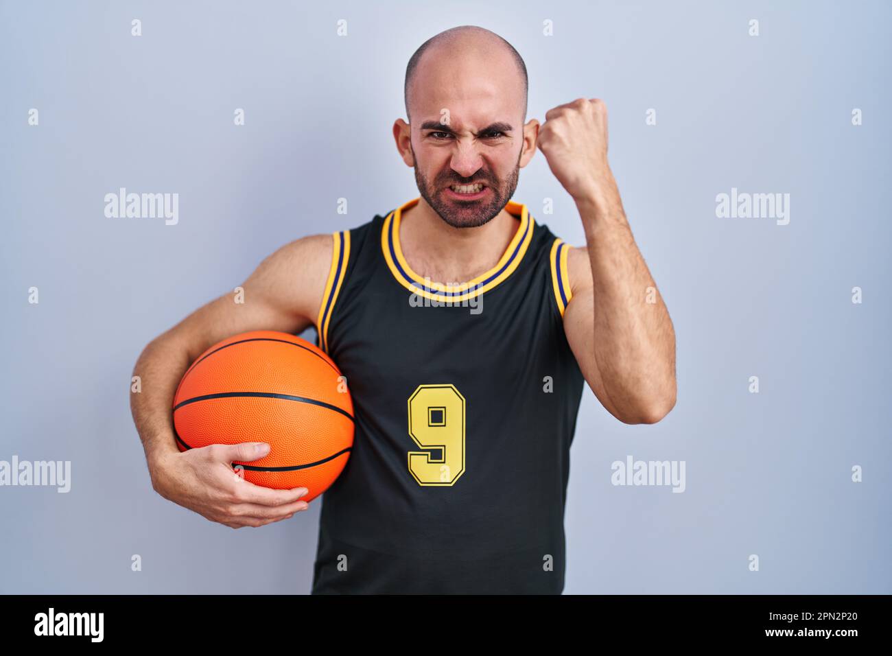Young bald man with beard wearing basketball uniform holding ball angry ...