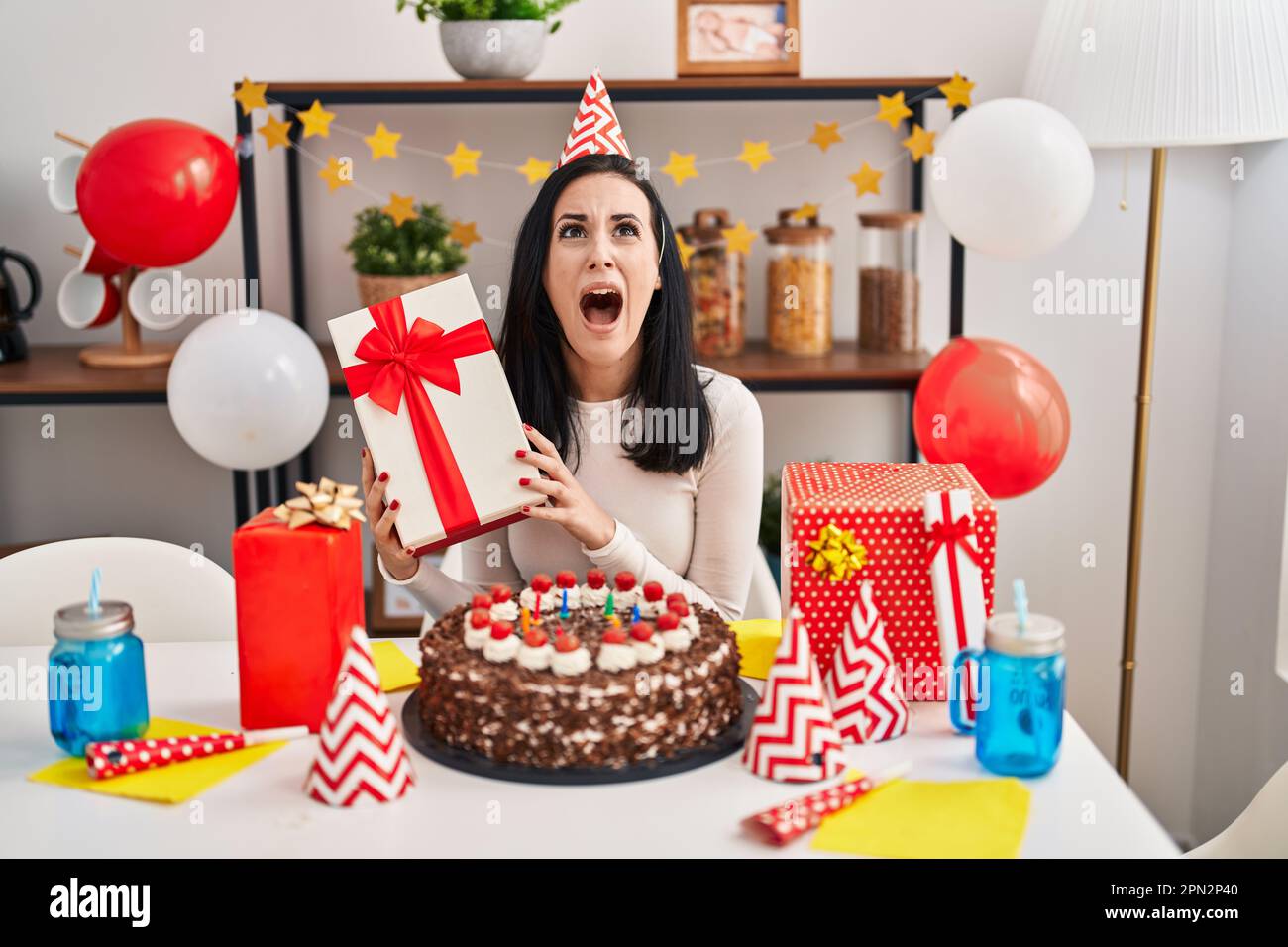 Hispanic woman celebrating birthday with cake holding present angry and ...