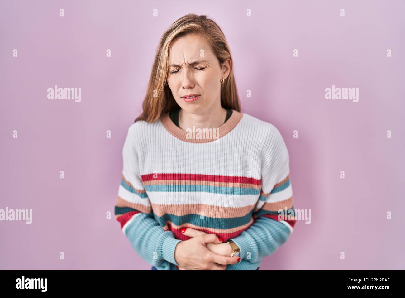 Young blonde woman standing over pink background with hand on stomach ...