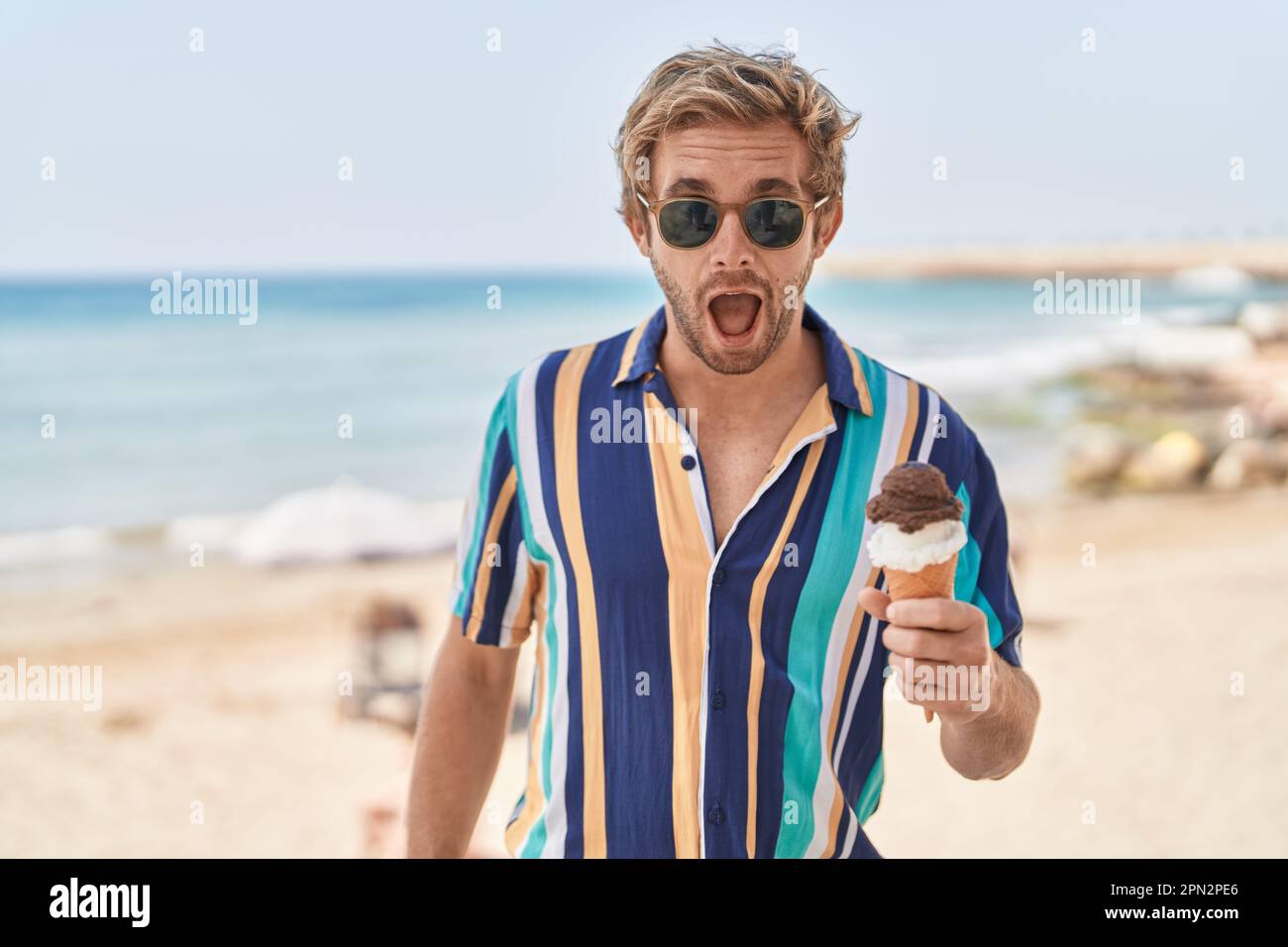 Caucasian man eating an ice cream at the beach scared and amazed with ...