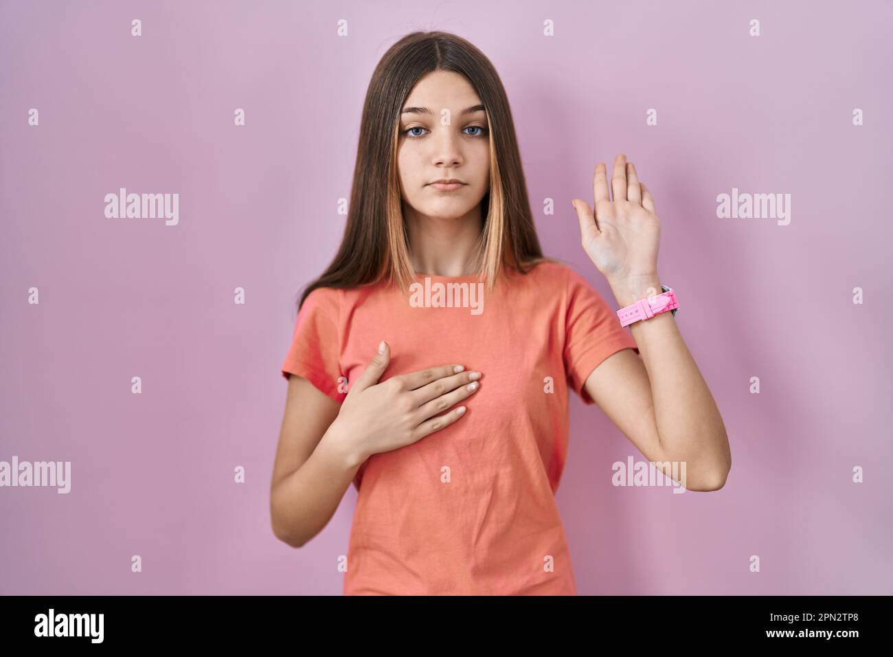 Teenager girl standing over pink background swearing with hand on chest ...