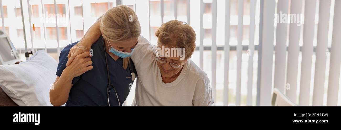 Female doctor in mask helping patient to get up from bed in the ward at ...