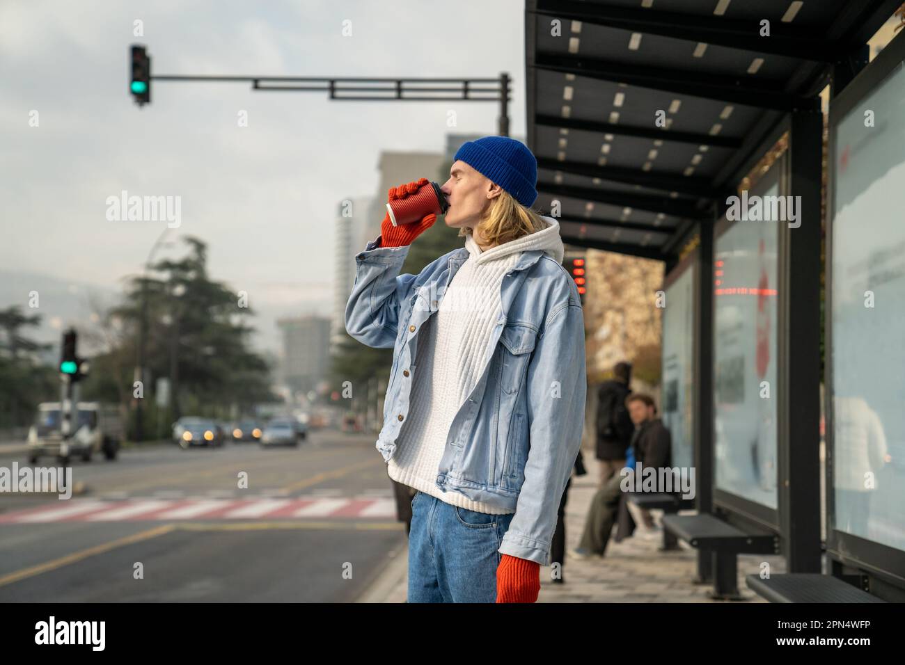 Stylish guy waiting bus standing on bus stop in downtown and drinking ...