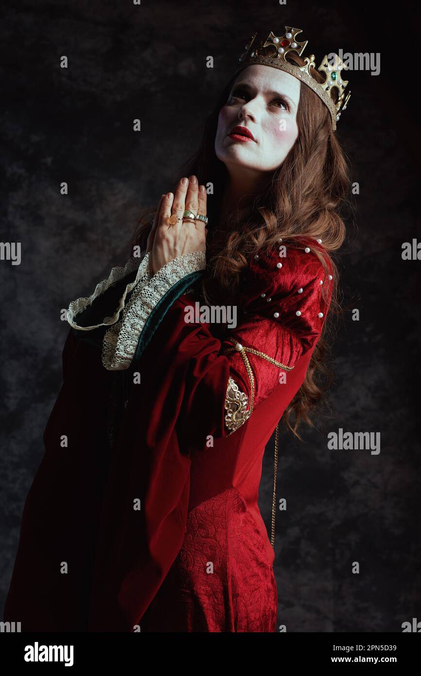 medieval queen in red dress with crown praying on dark gray background ...