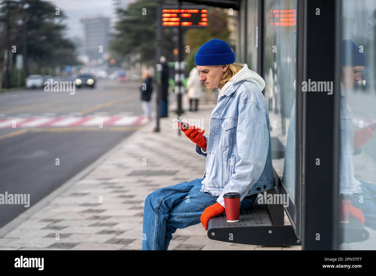 Stylish guy waiting bus sitting on bus stop bench in downtown with ...