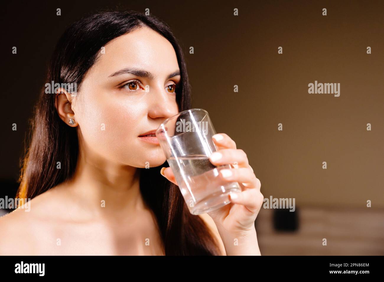 Pretty brunette drinking water standing near bed at home in the bedroom ...