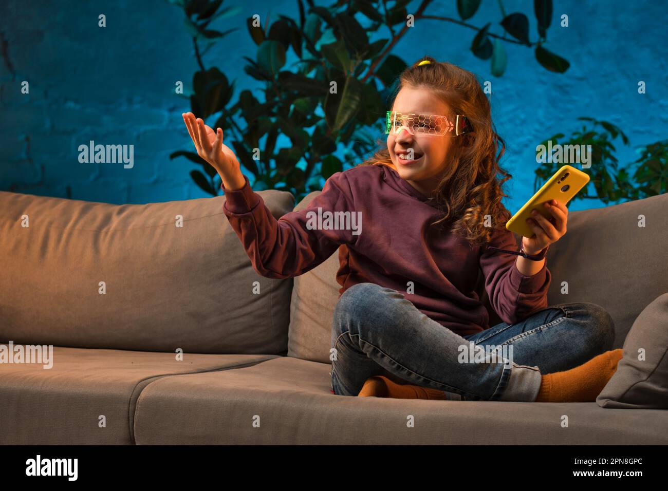 Teen girl in futuristic luminous glasses. Teenager sits on sofa and ...