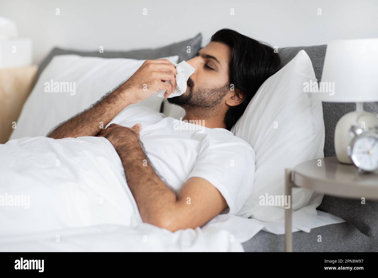 Sad young middle eastern guy in white t-shirt lies on bed, blows nose ...