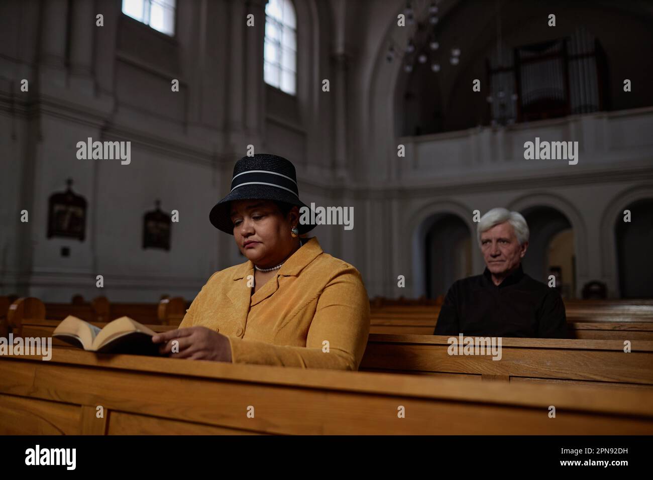 Elegant woman in hat reading Bible sitting on wooden bench in church ...