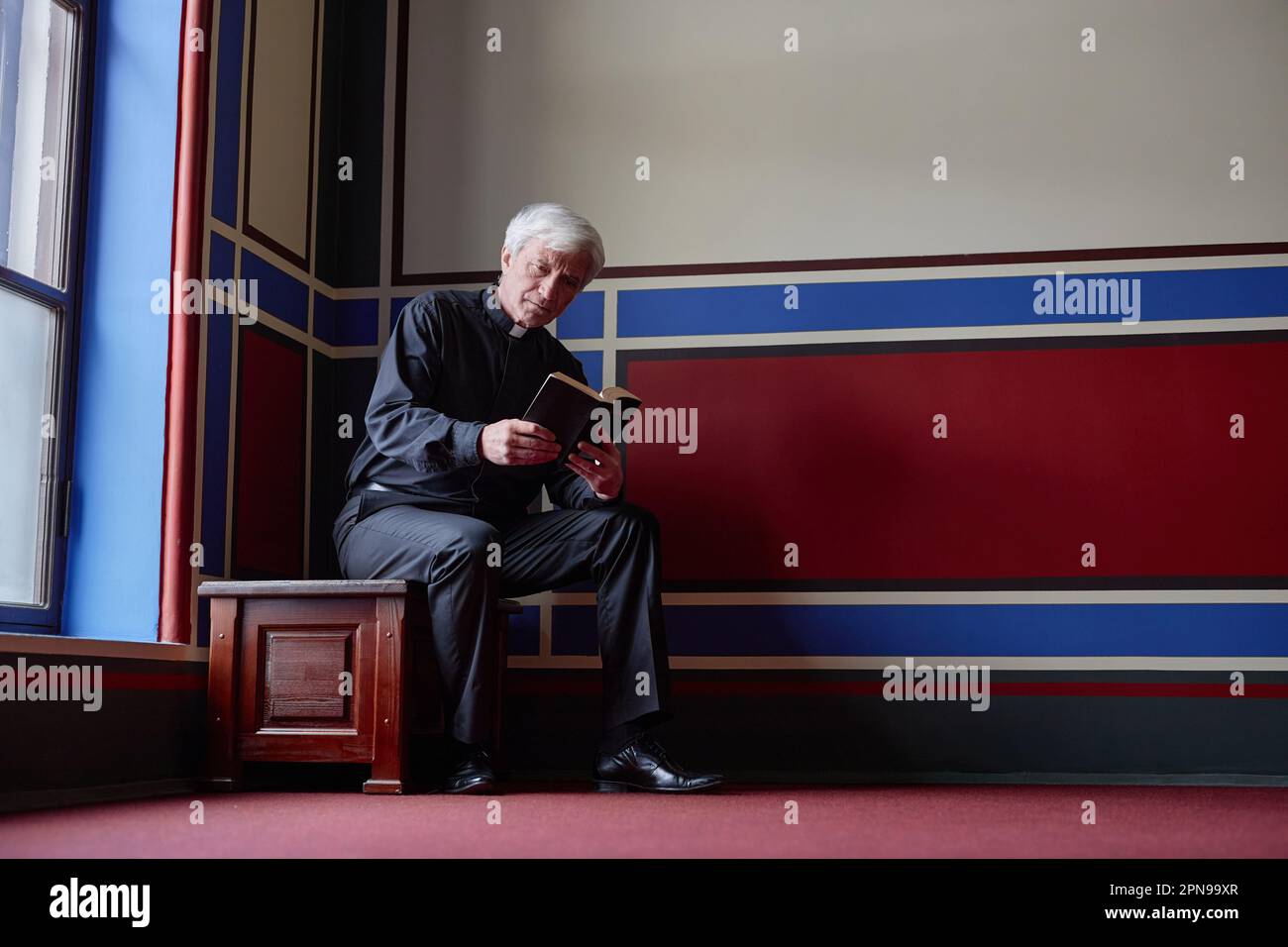 Senior priest reading Bible while resting on bench near the window in ...