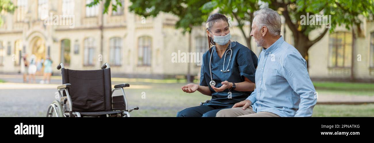 Talkative caring young nurse wearing face shield chatting with aged man ...