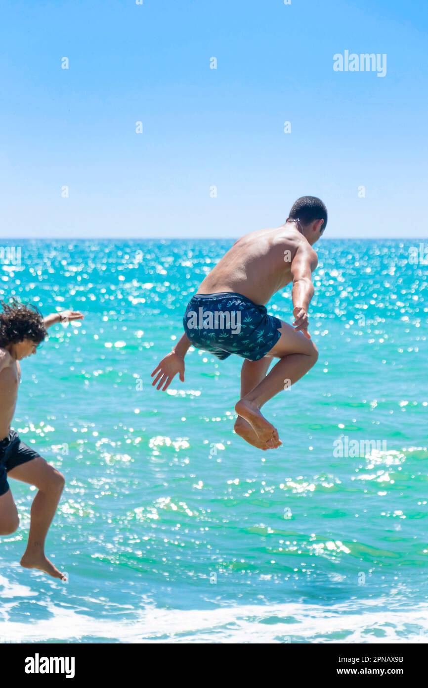 teenage boys in swimming shorts jumping into the sea from a pier in ...