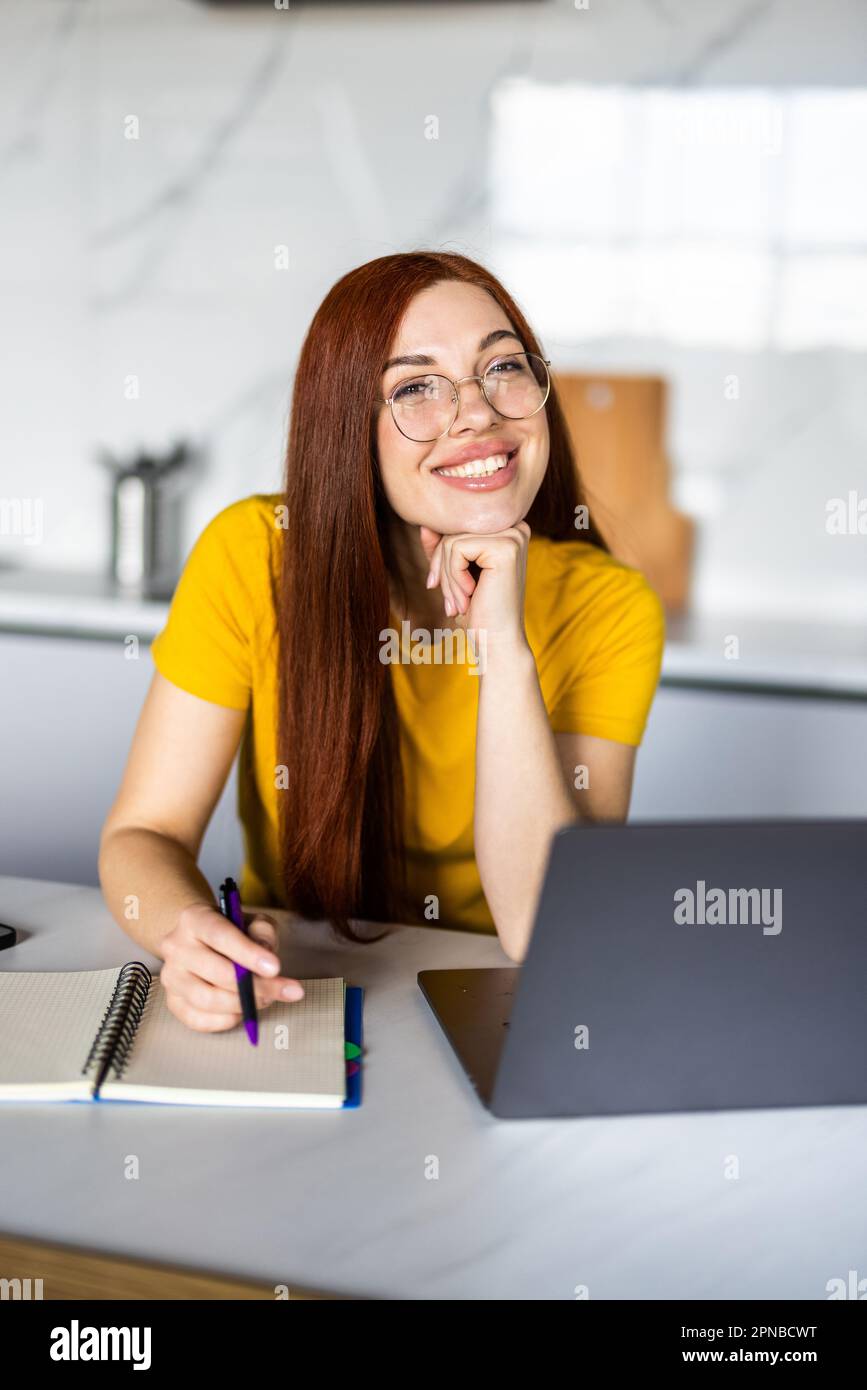 Young woman sit at table with laptop in kitchen holds pen, take notes ...