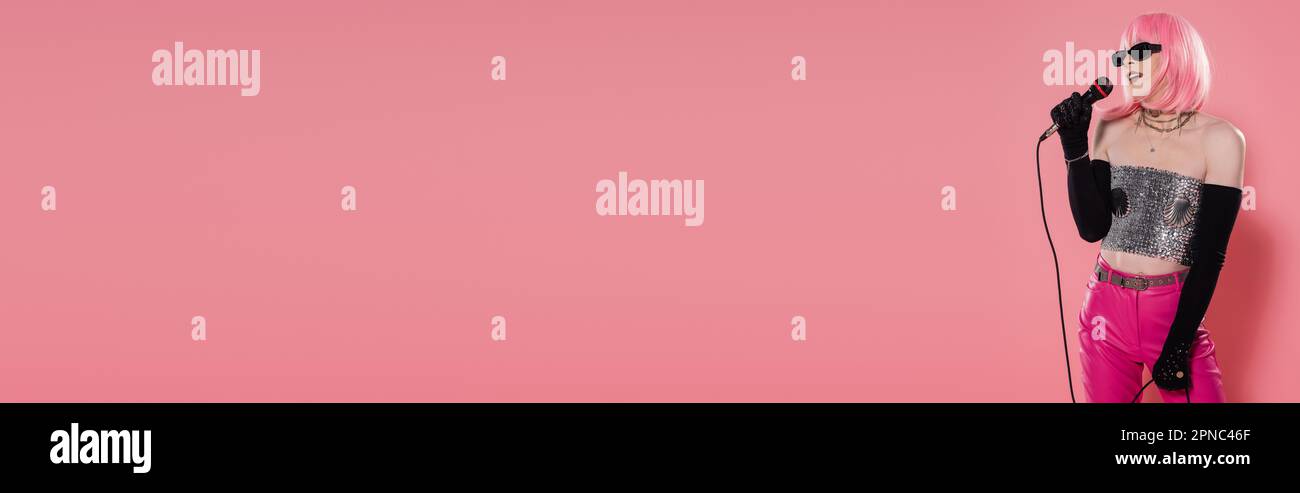 Stylish drag queen in sunglasses and gloves holding microphone on pink ...