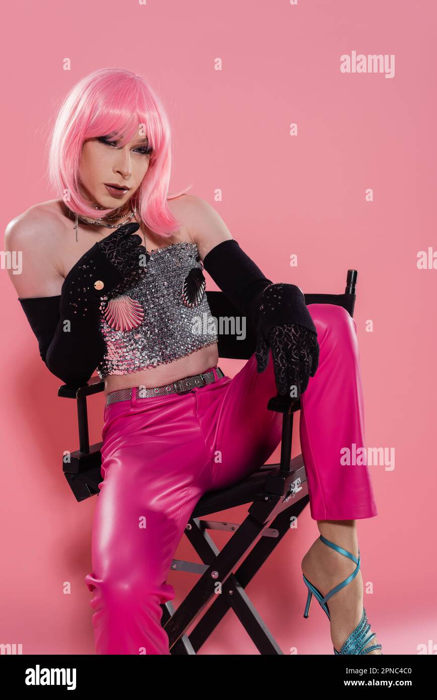 Fashionable drag queen in gloves and top sitting on chair on pink ...