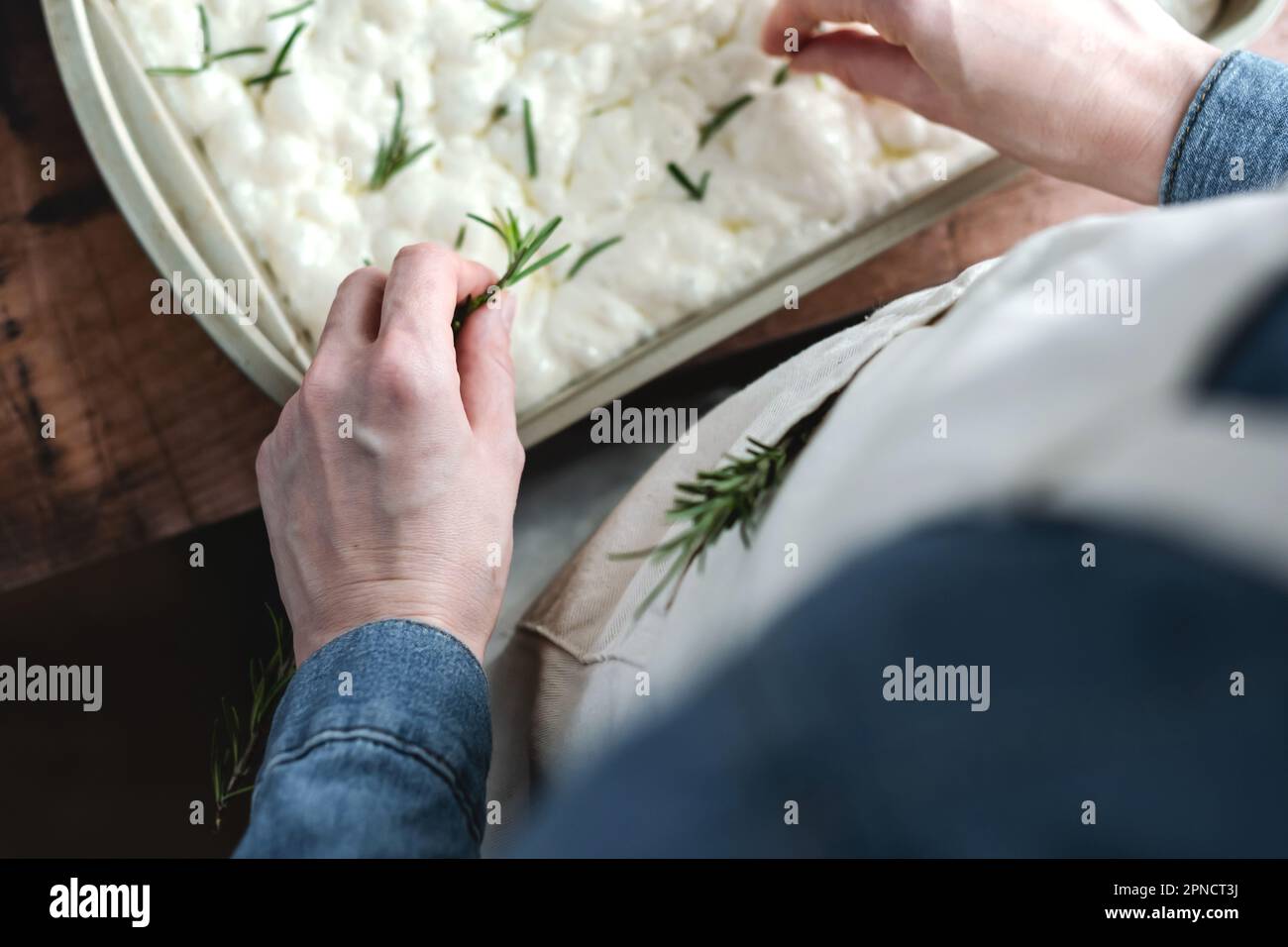 Female hands pinching rosemary leaves and topped on dough focaccia. The ...