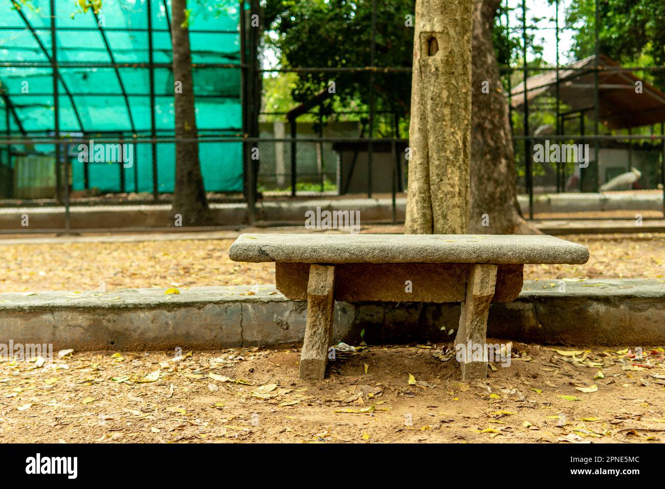 a park bench made of stone with a tree in the background. has a green net-covered shaded cage on ...