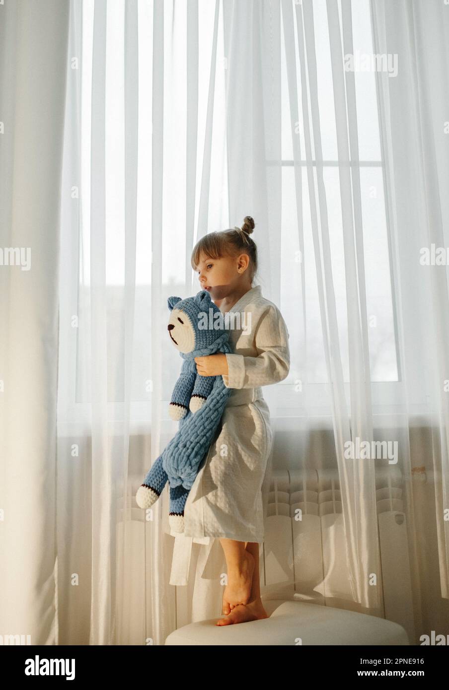 A girl in a dressing gown stands by the window and plays with a knitted ...