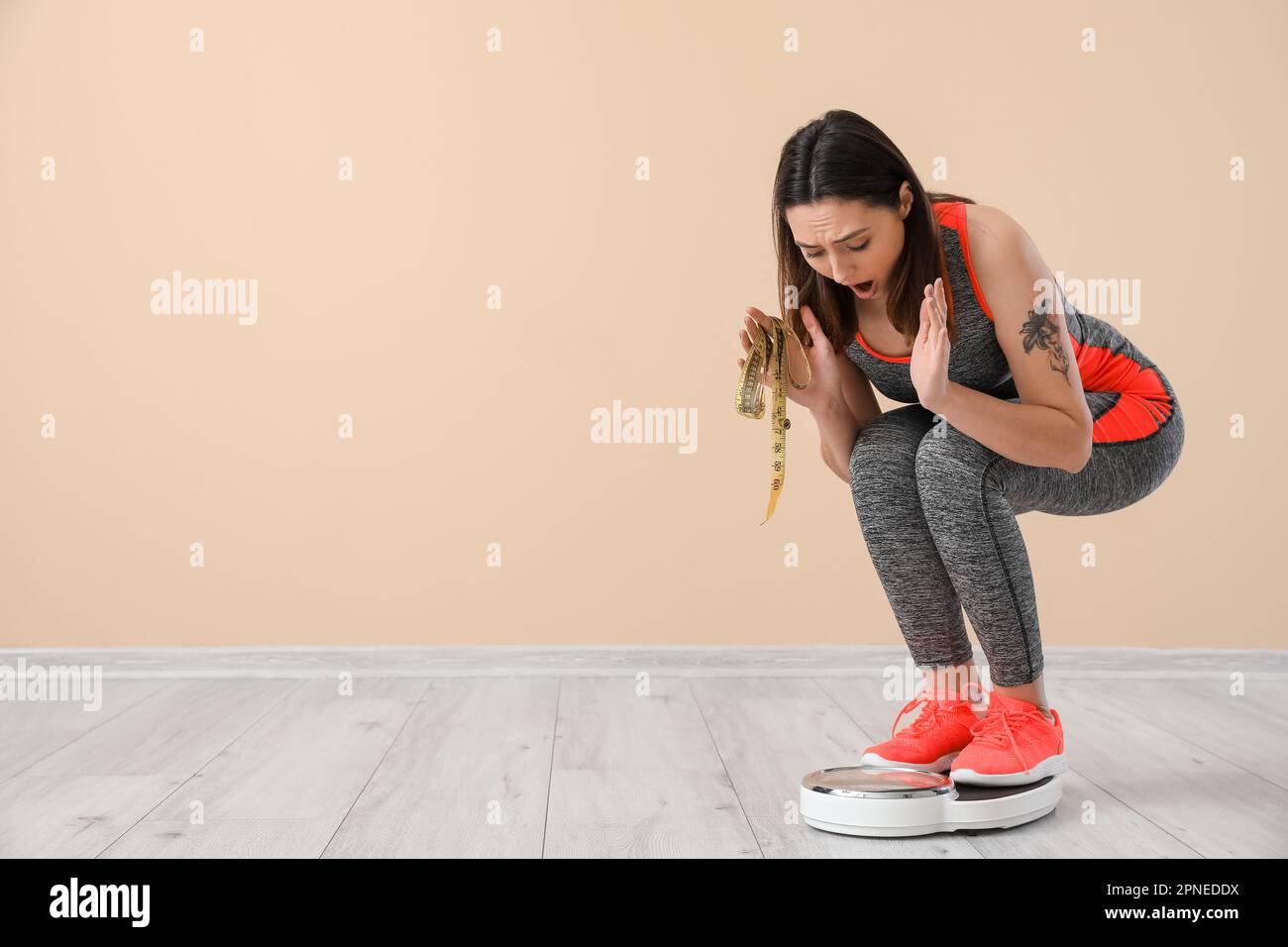 Shocked young woman measuring her weight on scales near beige wall ...