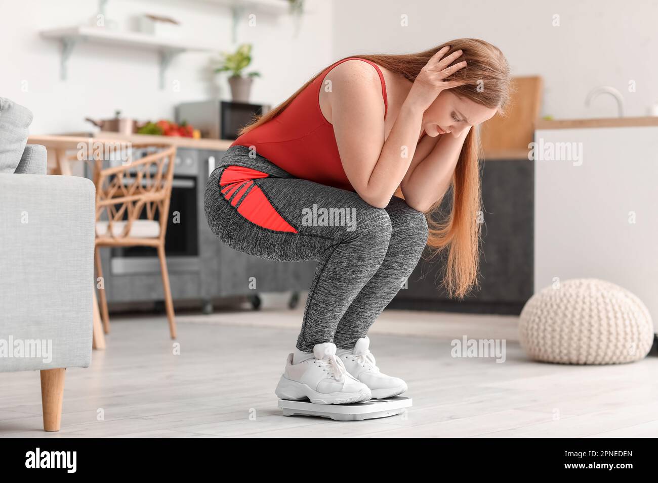 Upset young overweight woman measuring her weight on scales in kitchen ...
