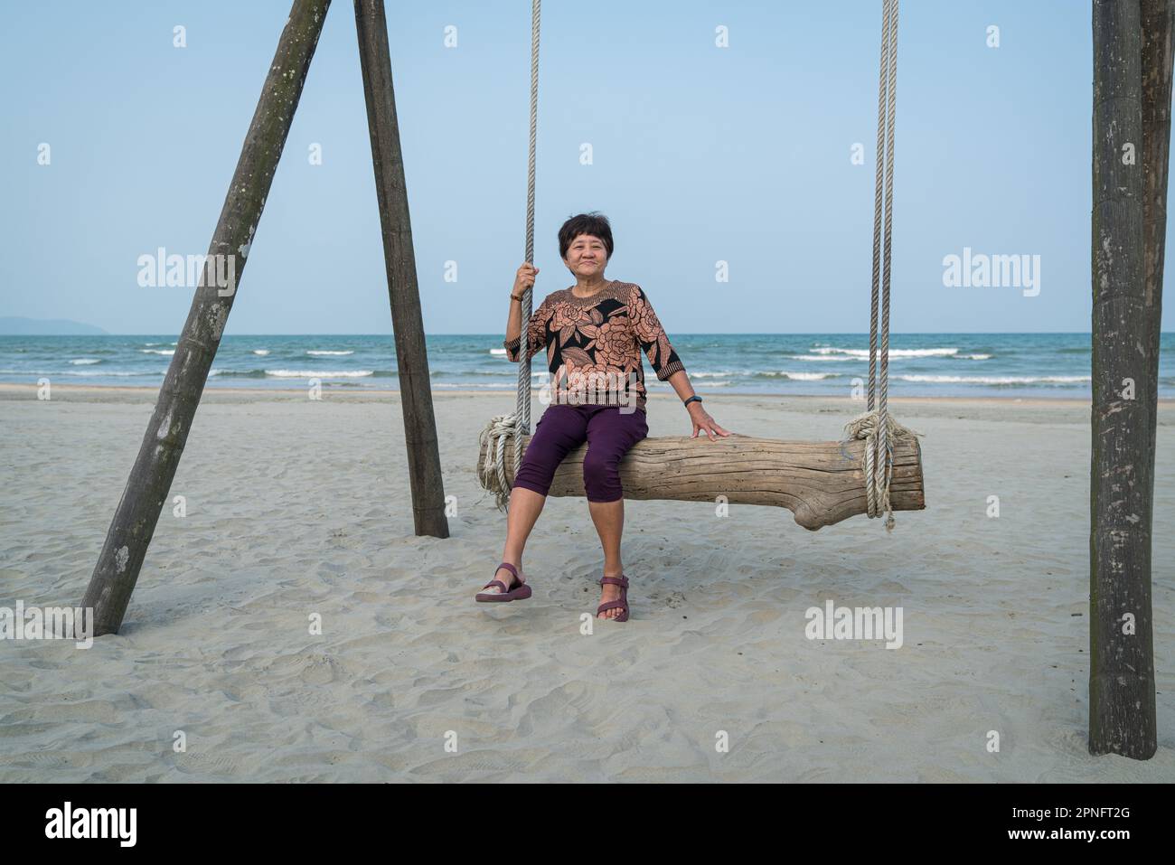 Senior Asian woman relaxing on a wood plank swing on the beach ...