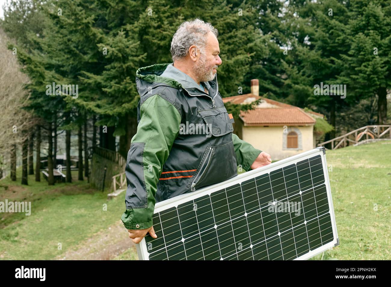 Middle aged bearded man carrying solar panel near house in rural zone ...