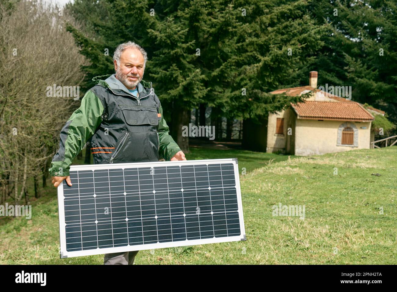 Middle aged bearded man carrying solar panel near house in rural zone ...