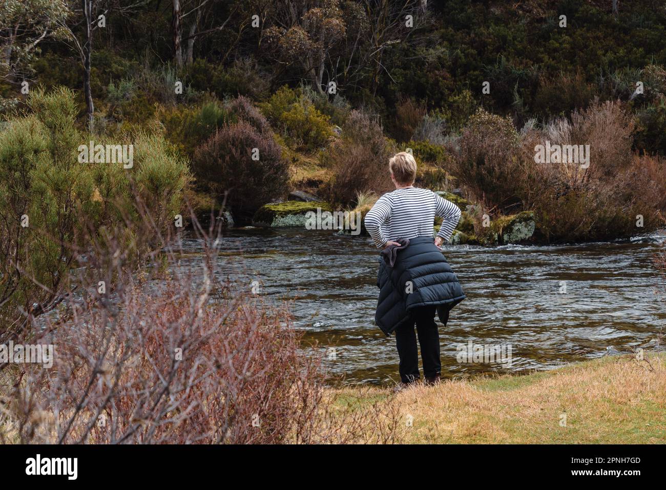 A back view of a female standing at the edge of a river, wearing a coat ...